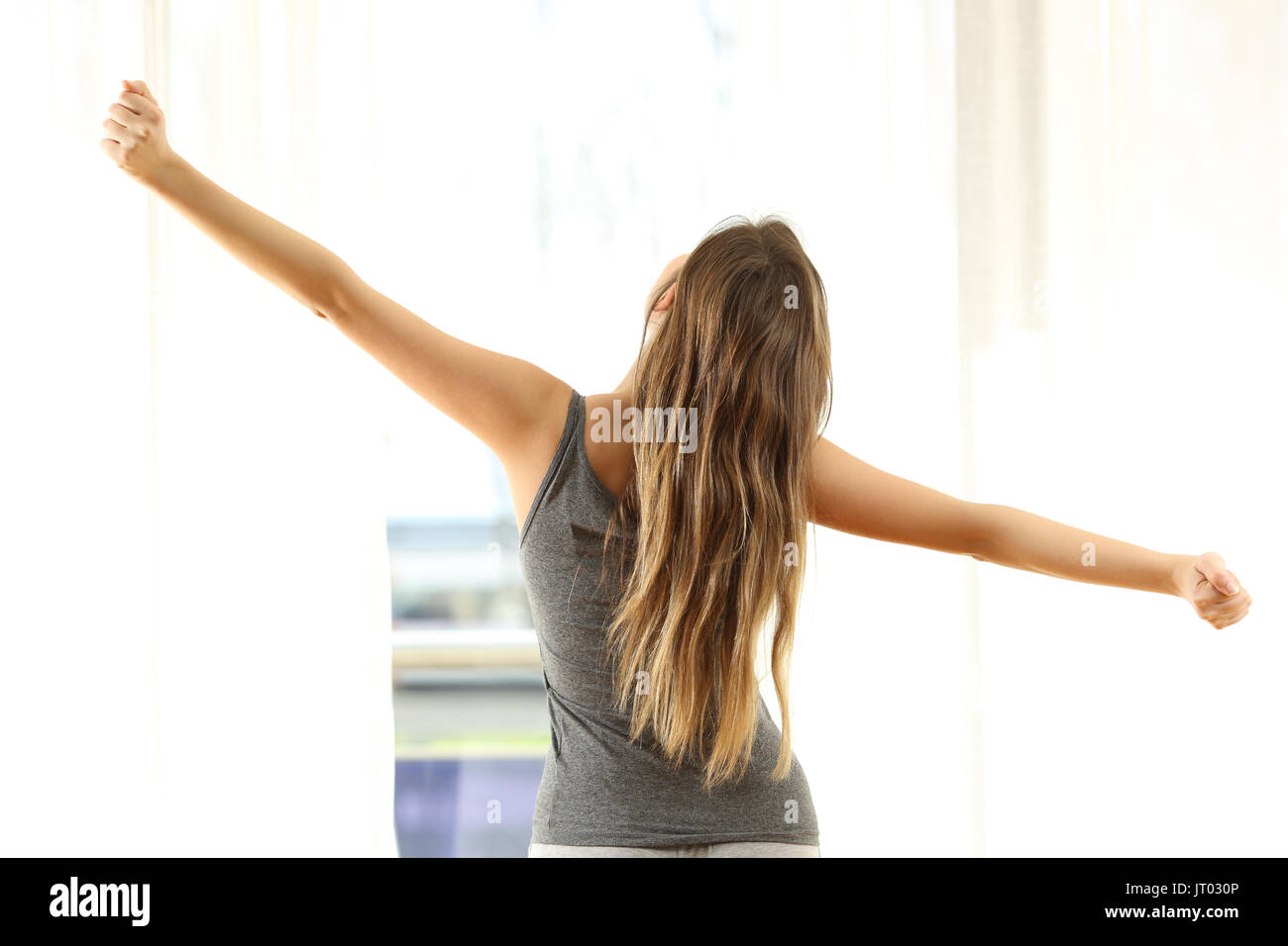 Back view of a girl stretching arms waking up looking through window in ...
