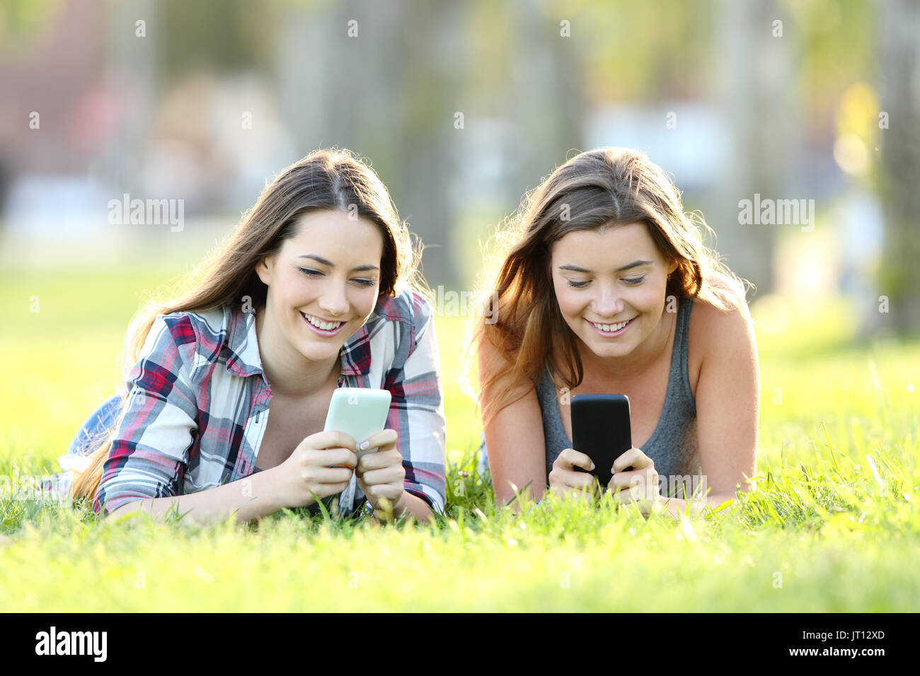 Front view of two happy friends using their smart phones on the grass ...
