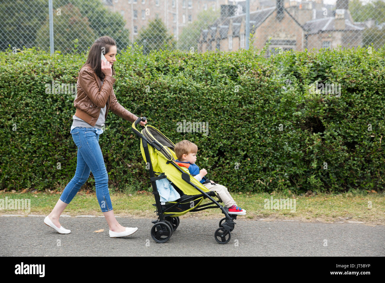 Busy Mum talking on phone while pushing her toddler sitting in a pram ...