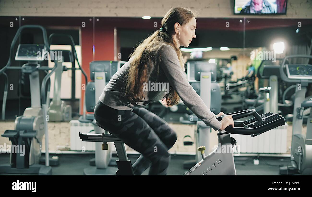 Young beautiful girl in gym, shaking her legs on cycling simulator ...