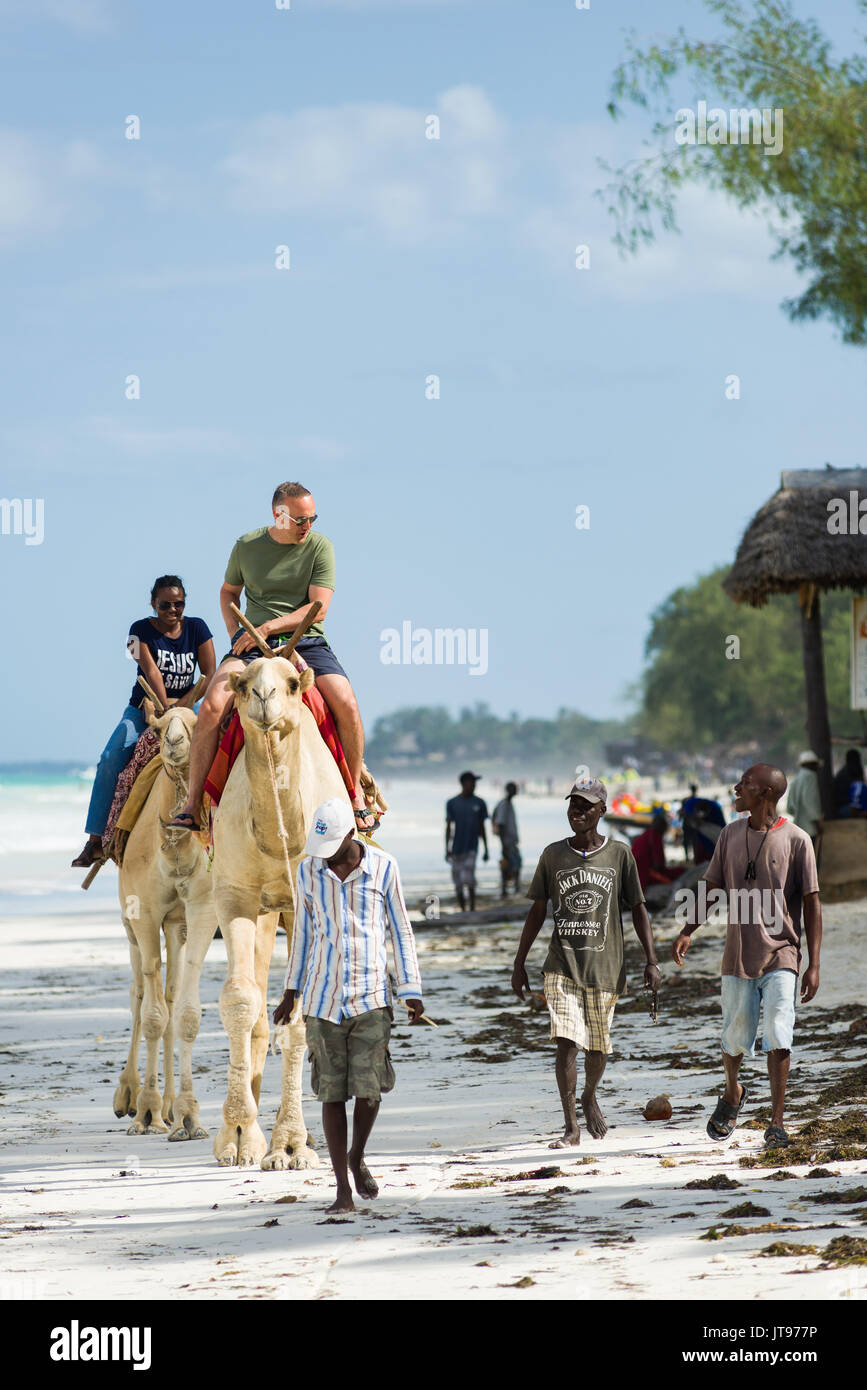 Two tourists riding on camels on the beach with locals walking along ...