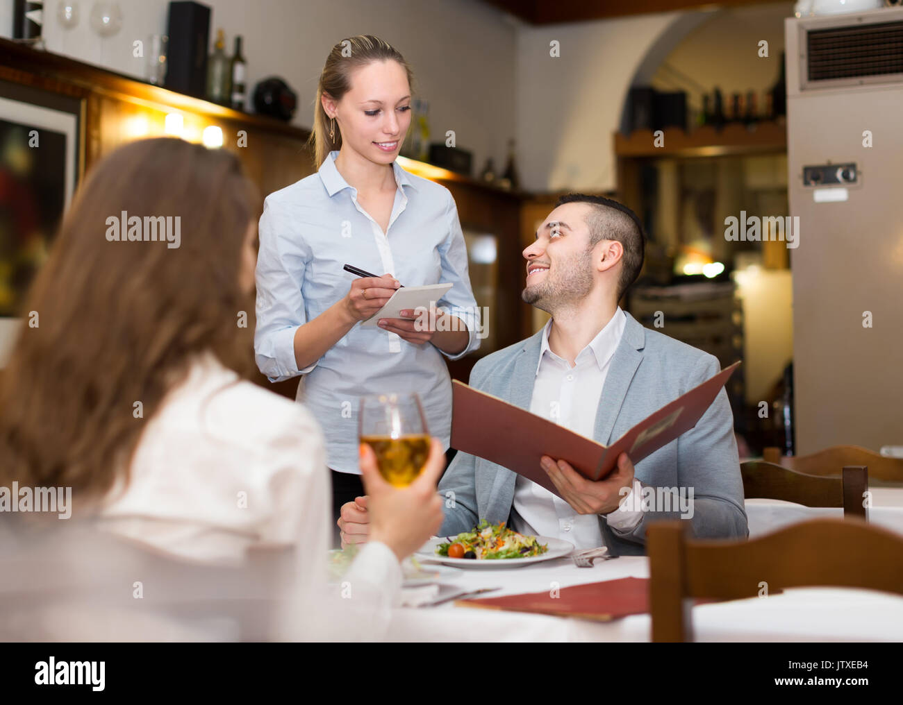 Young blonde waitress offering tasty dishes to smiling guests couple ...
