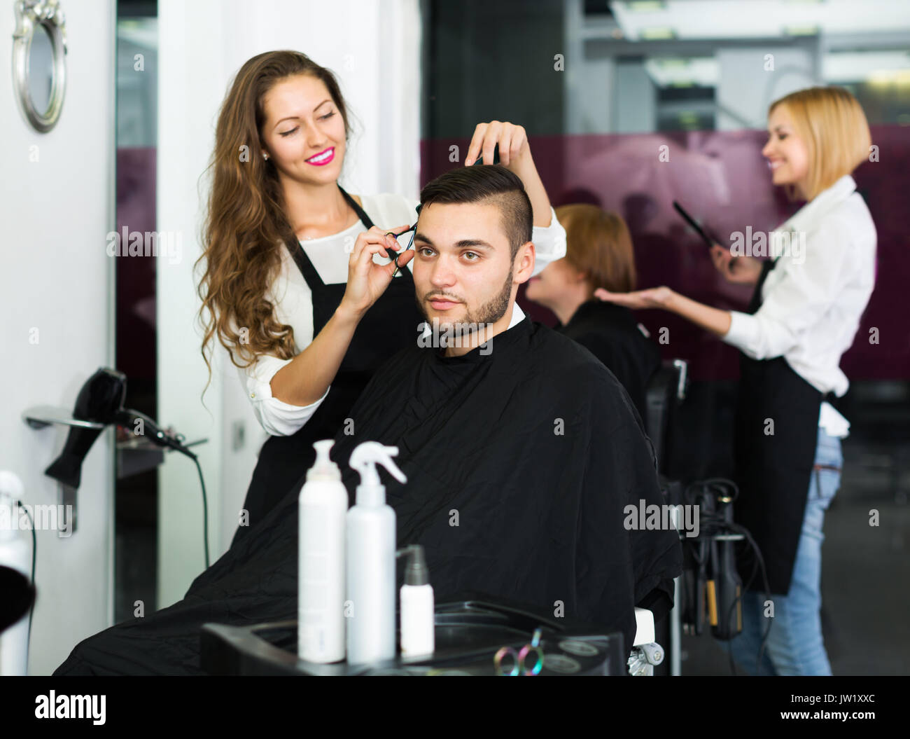 Happy professional young hairdresser doing hairstyle for young men ...