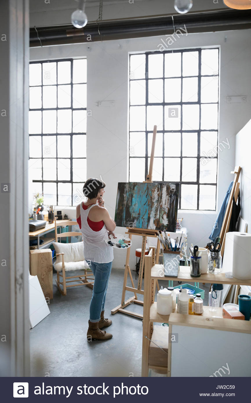 Female painter with palette examining canvas painting on easel in art ...