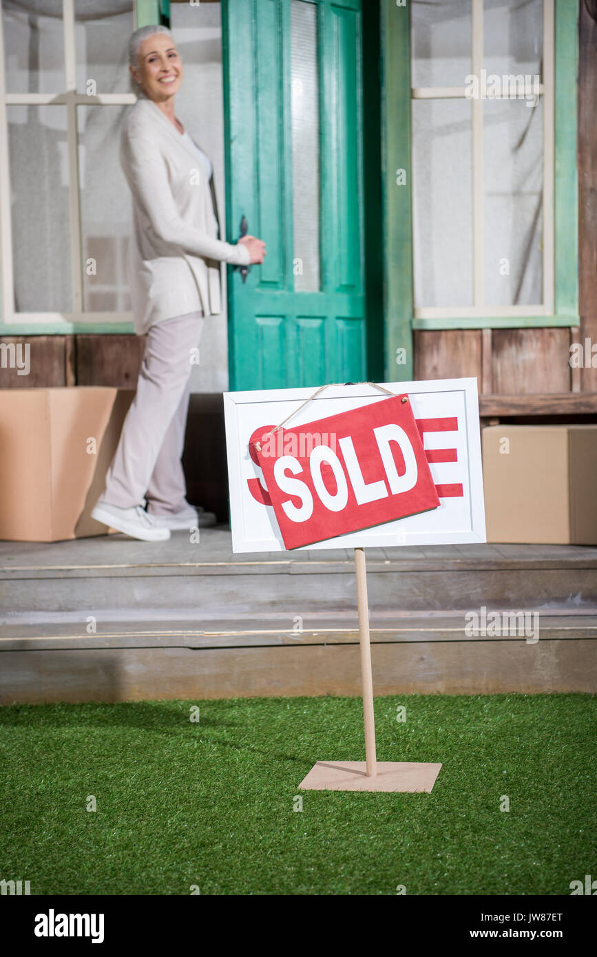Smiling senior woman standing on porch of new house with sold sign ...