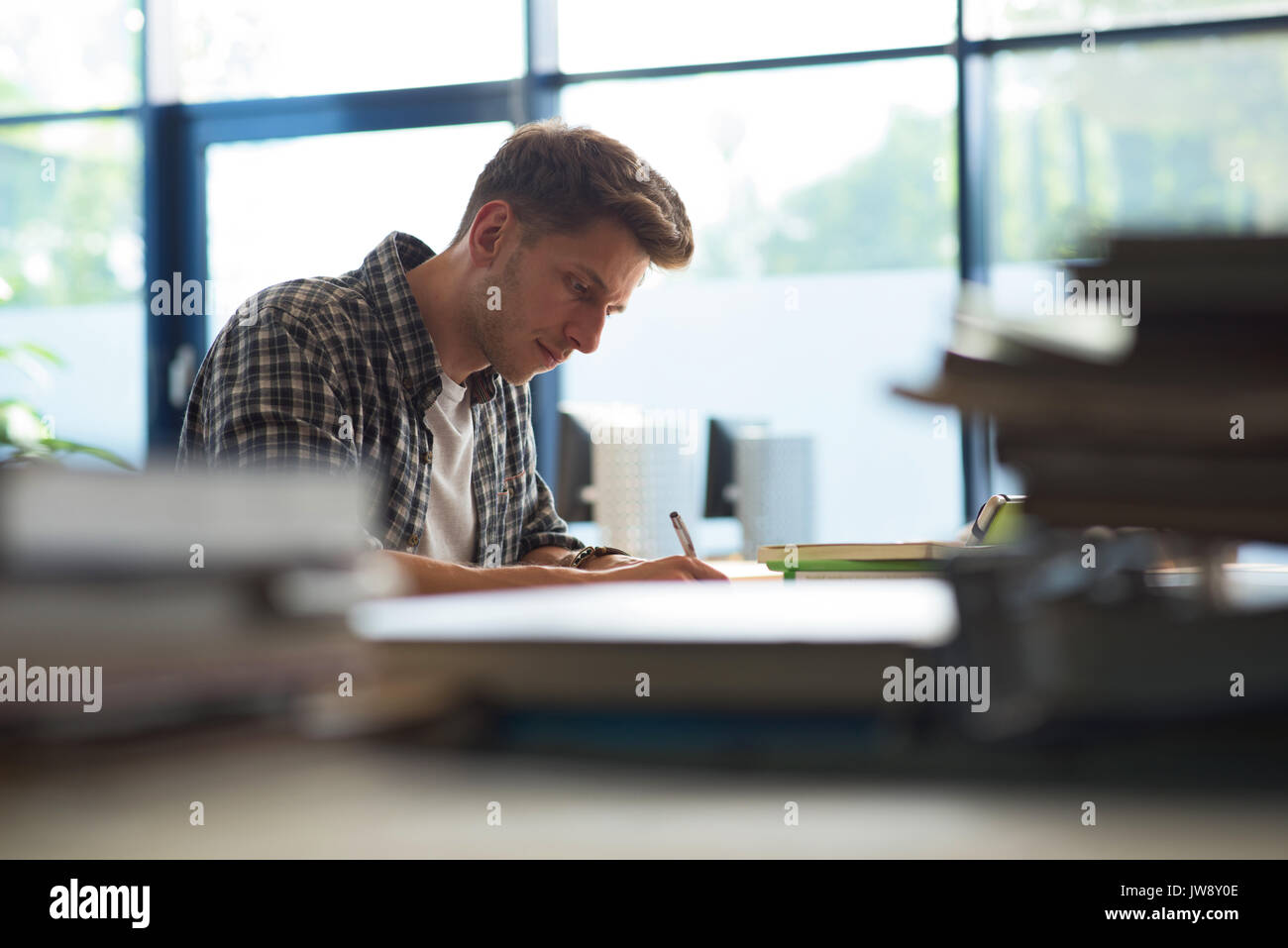 Side view of young male student studying at desk by window in classroom ...