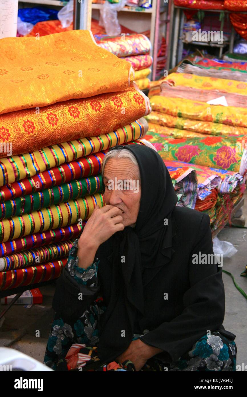 Old lady sitting in front of bales of colourful silk at the Kashgar ...