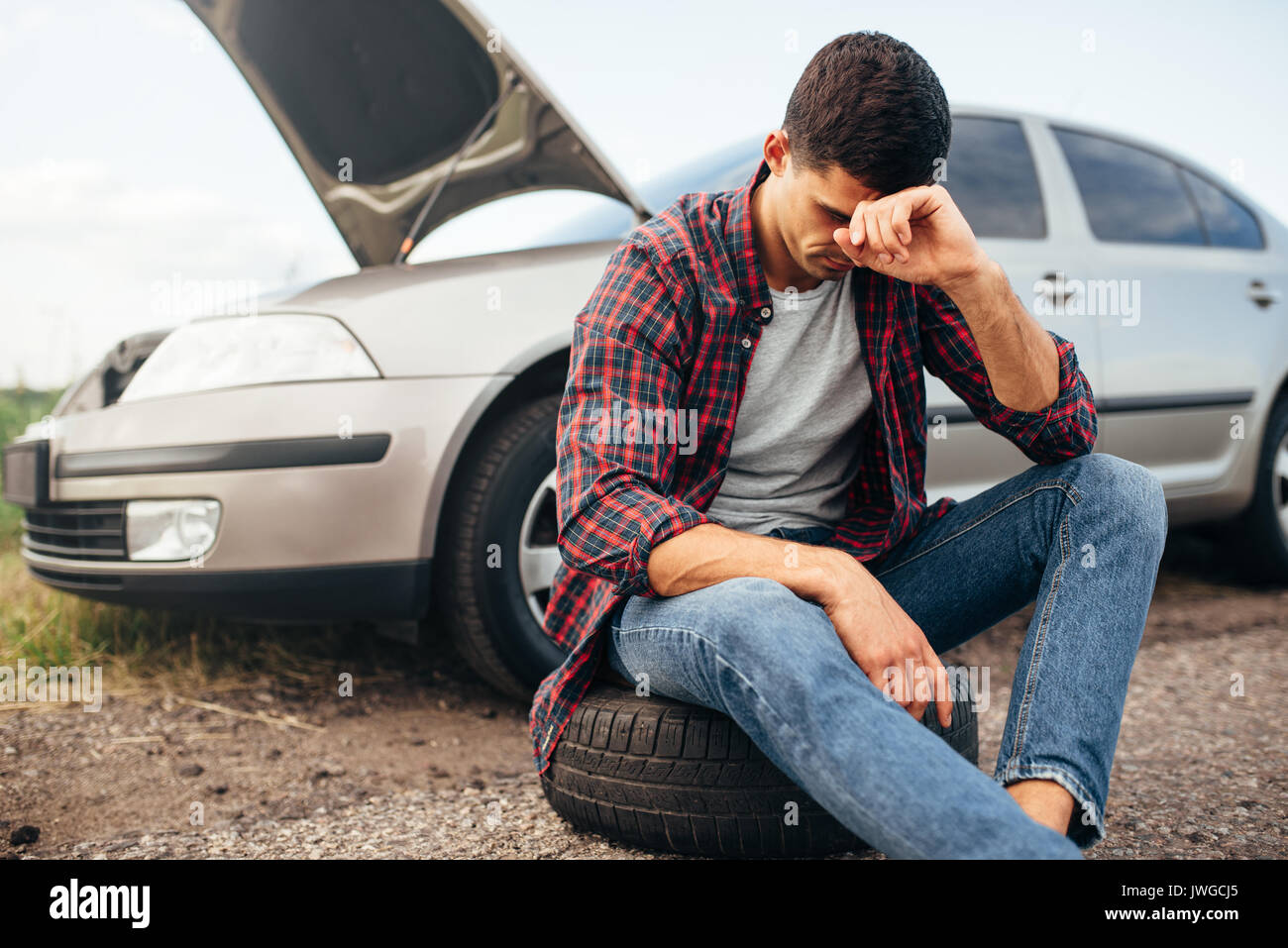 Tired man sitting on tire, broken car with open hood on background ...