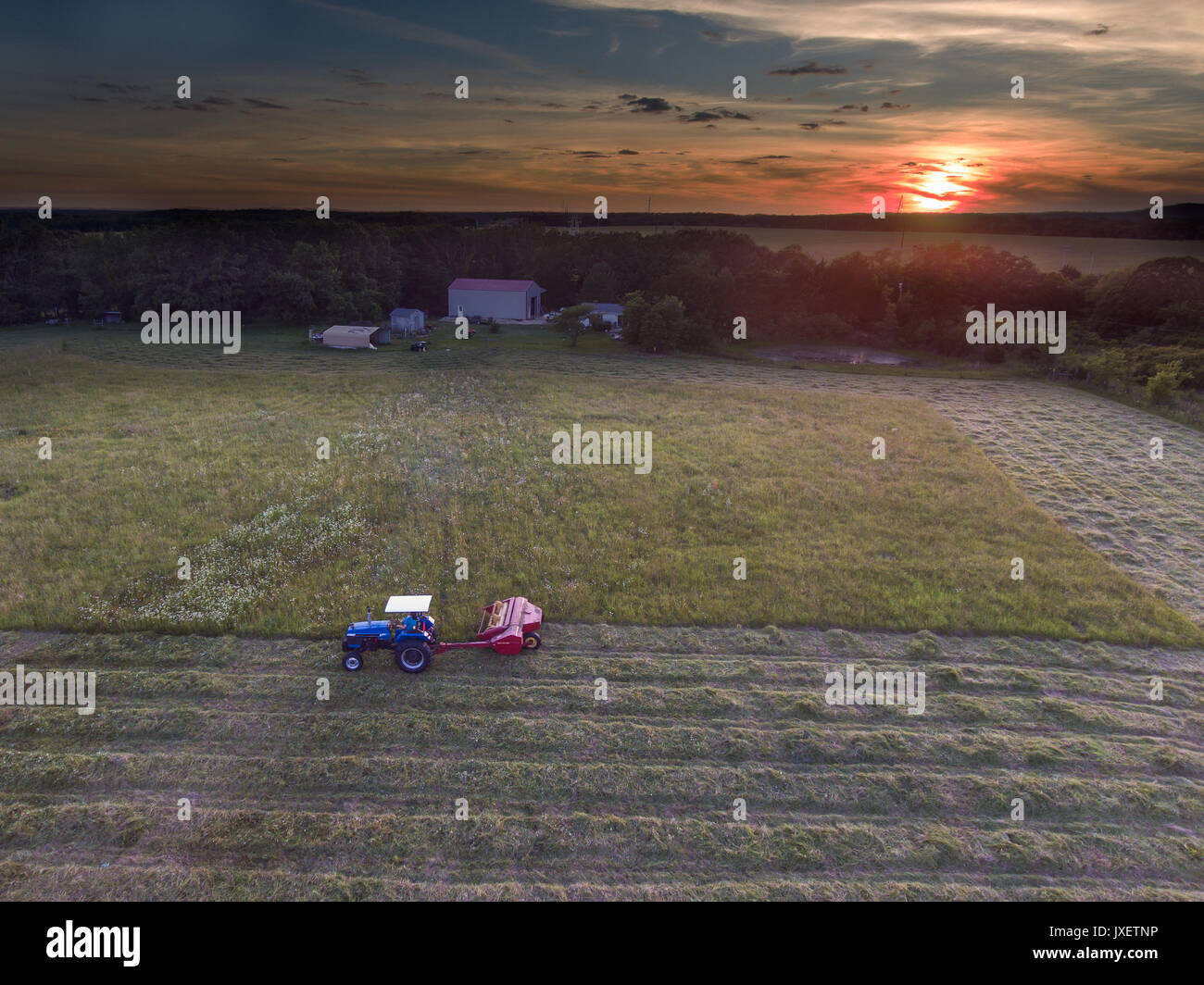 Blue tractor pulling a hay cutter in small field with farm and sunset ...