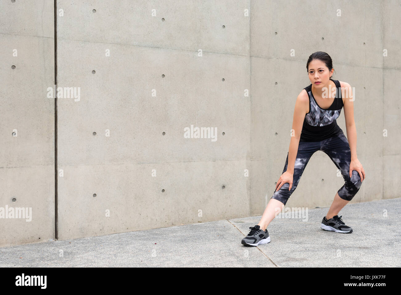 young woman standing on outside gray wall background making legs warmup ...