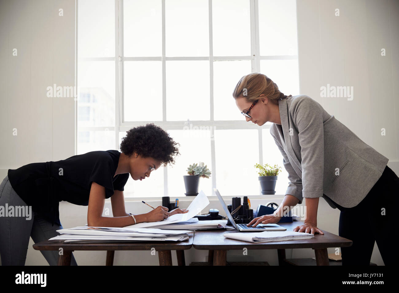 Two Women Work Leaning Over Opposite Sides Of An Office Desk Stock