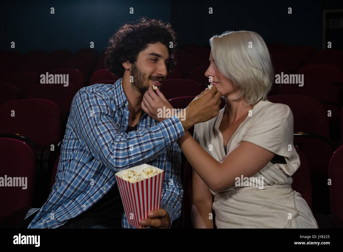Happy couple feeding popcorn to each other while sitting in theatre ...