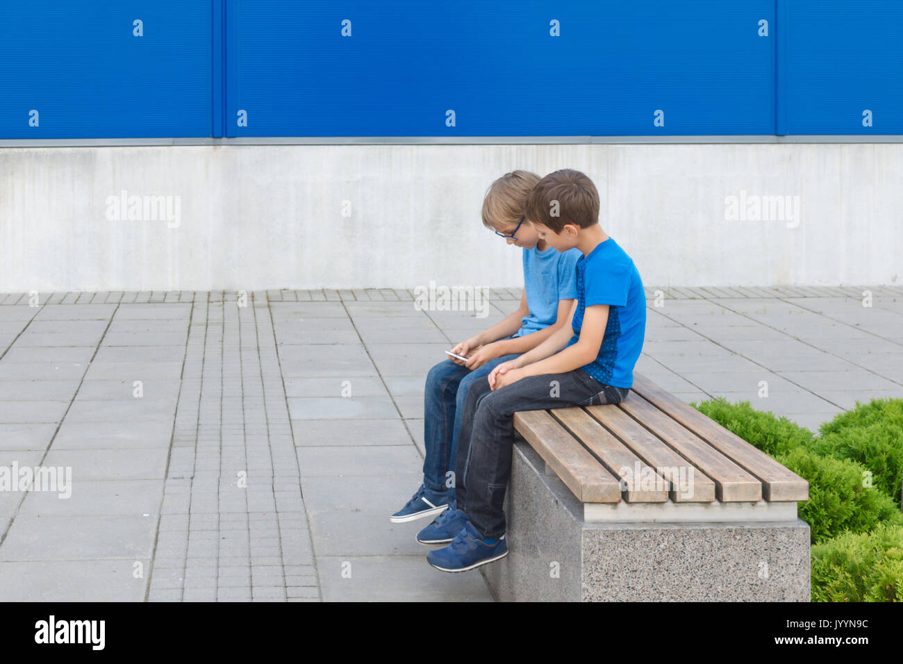 Two little boys playing with mobile phone sitting on the bench outdoors ...