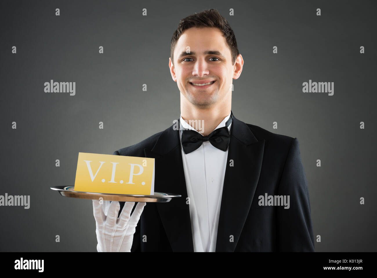 Portrait of happy young waiter holding tray with VIP sign against gray ...