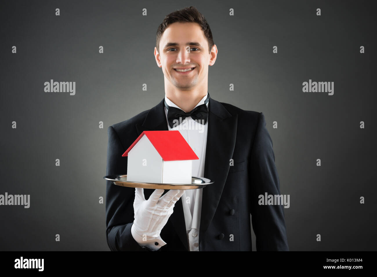 Portrait of happy young waiter holding house model in tray against gray ...