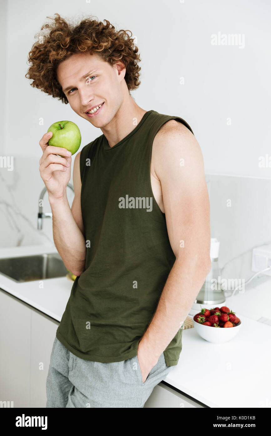Picture of curly smiling man standing at the kitchen and holding apple ...