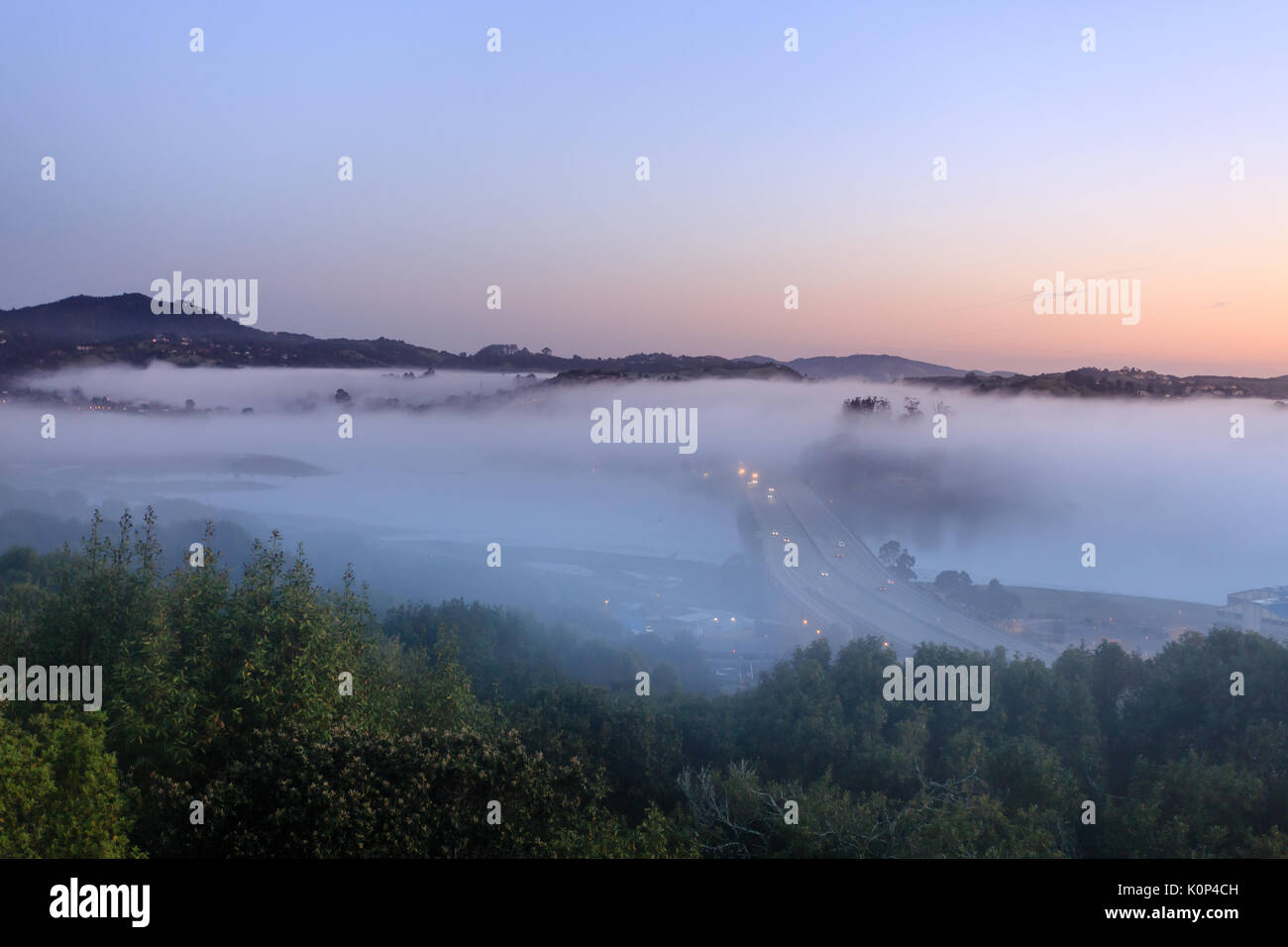 Sunrise reveals low mist and fog over bay and roadway lights on highway ...
