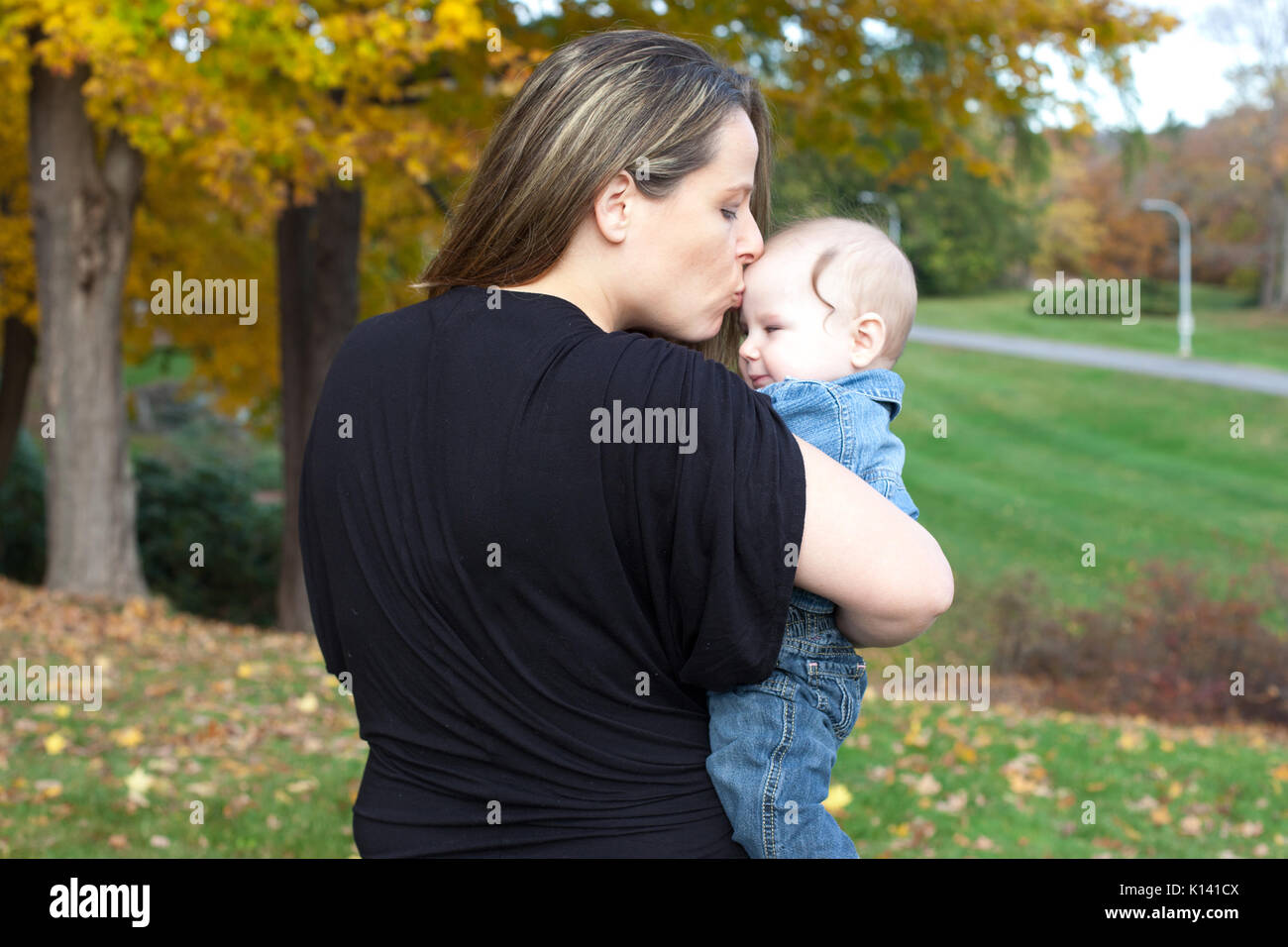 Rear view of a young woman in a park kissing her baby daughter on her ...