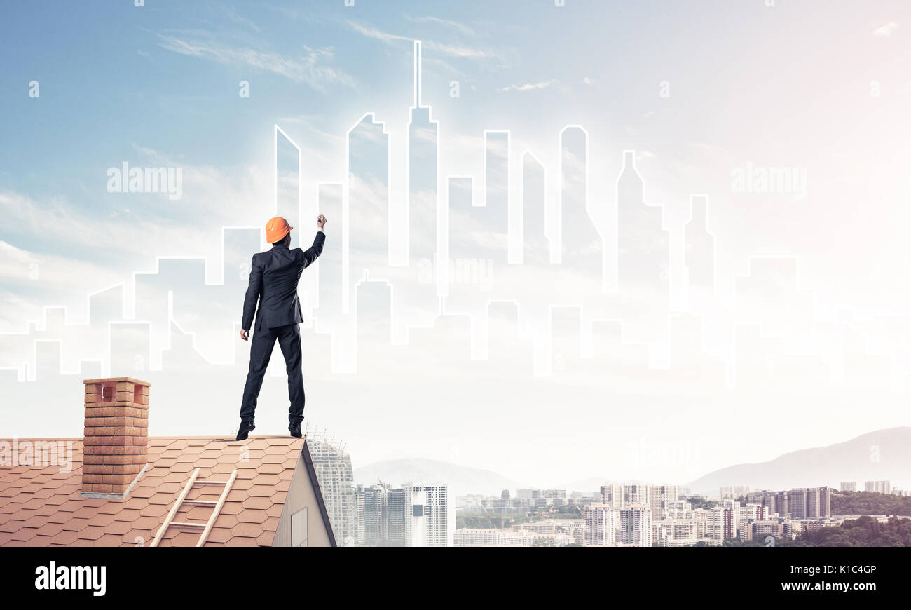 Engineer man standing with back on house roof and drawing city. Mixed ...