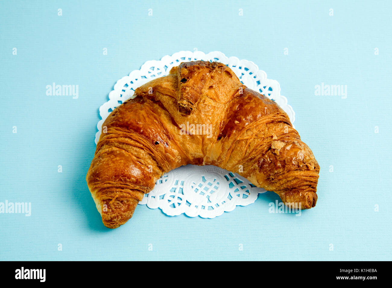 a french croissant on a doily lace paper. Pastel blue background ...