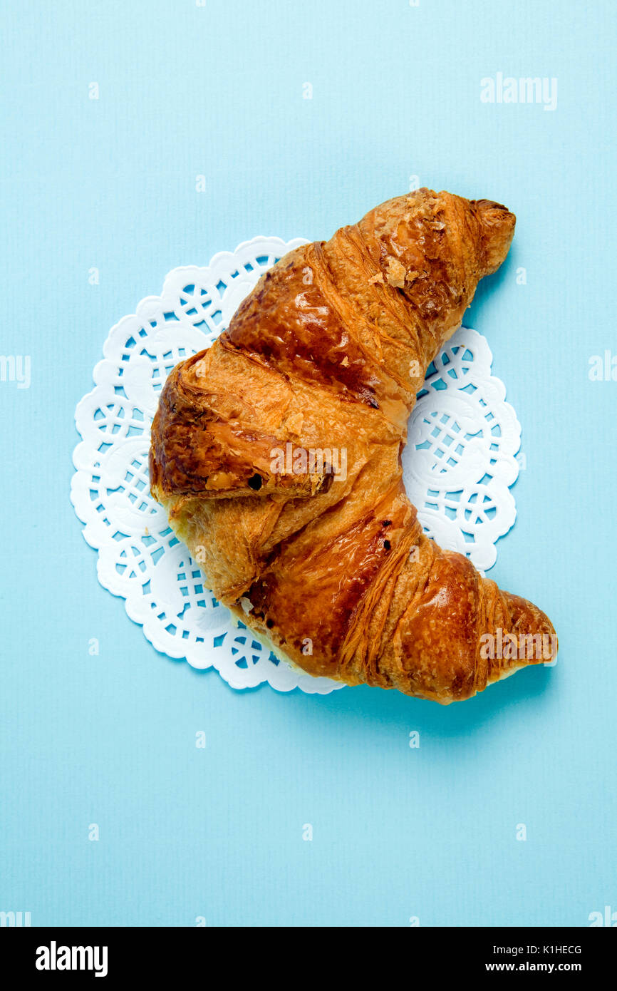 a french croissant on a doily lace paper. Pastel blue background ...