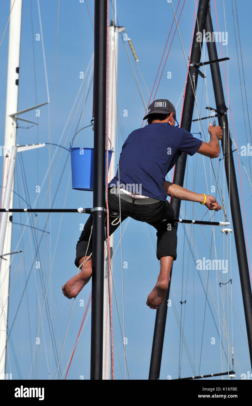 A man aloft up a mast on a large yacht or sailing boat in cowes yacht ...
