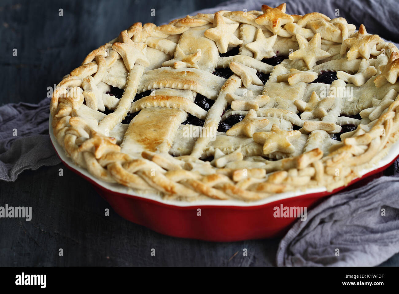 Moody shot of a blueberry pie with lattice and stars crust in a red pie ...