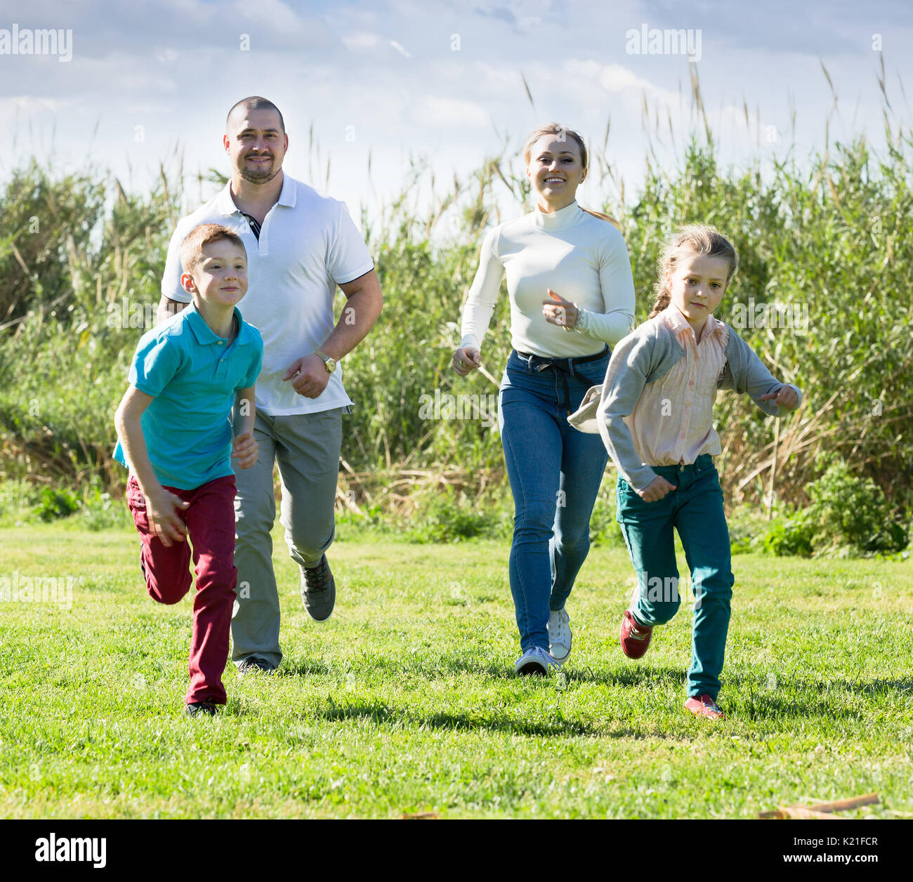 Joyful young parents with two kids playfully running on the green lawn ...