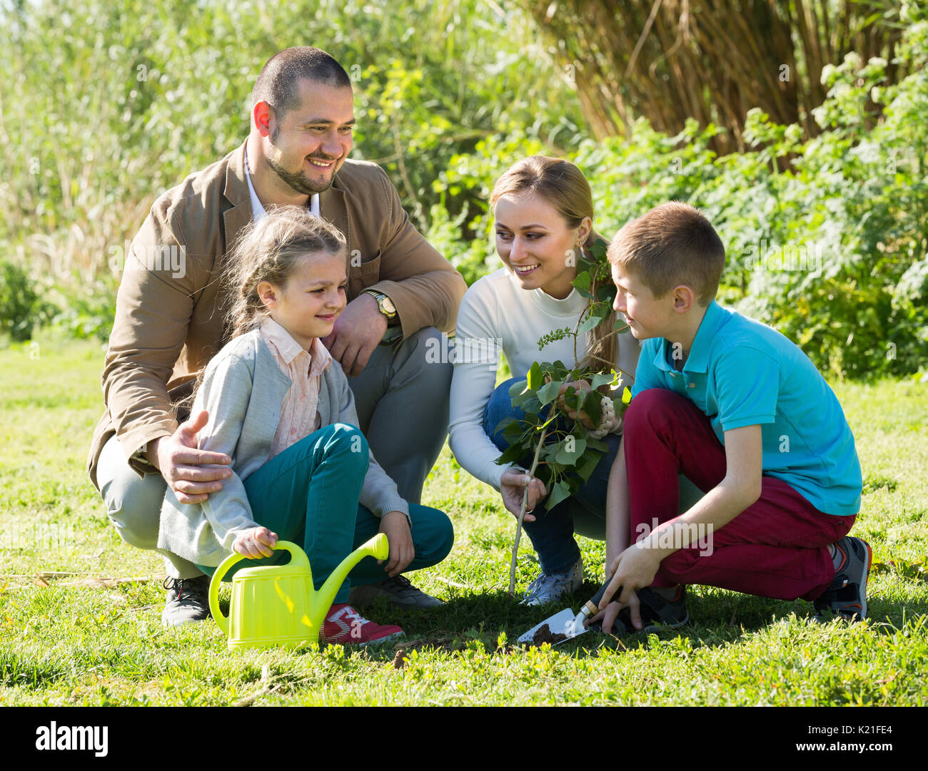 Cheerful young parents with two smiling kids planting a tree together ...