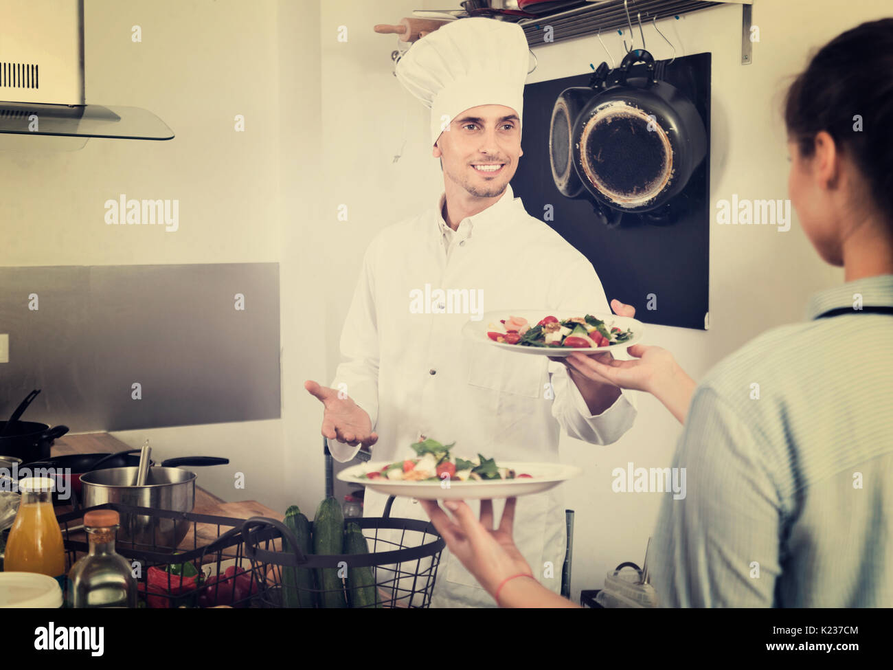 Portrait of diligent young man cook giving to waitress ready to serve ...