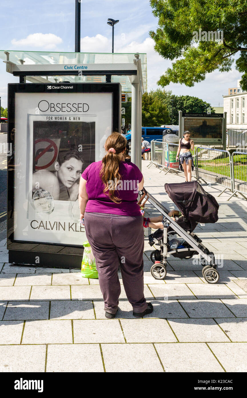 A large woman stands in front of a bus stop featuring an advertisement ...