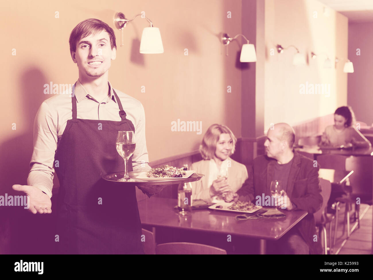 Professional smiling waiter with tray posing at table of senior ...