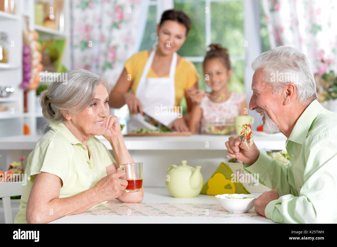 Senior couple drinking tea at kitchen, mother and daughter cooking on ...