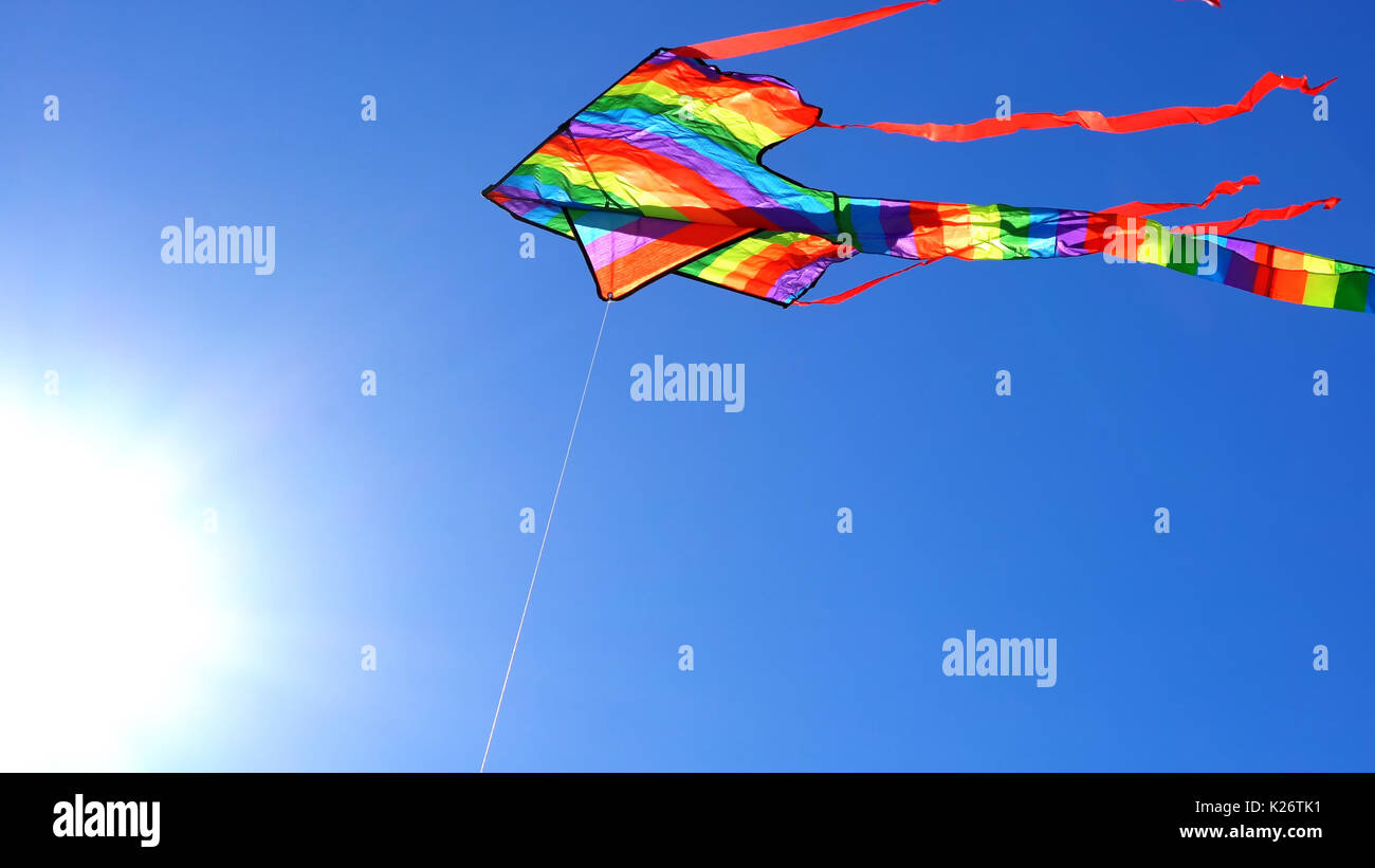 Colorful rainbow kite flying against a sunny blue sky, with lens flare ...