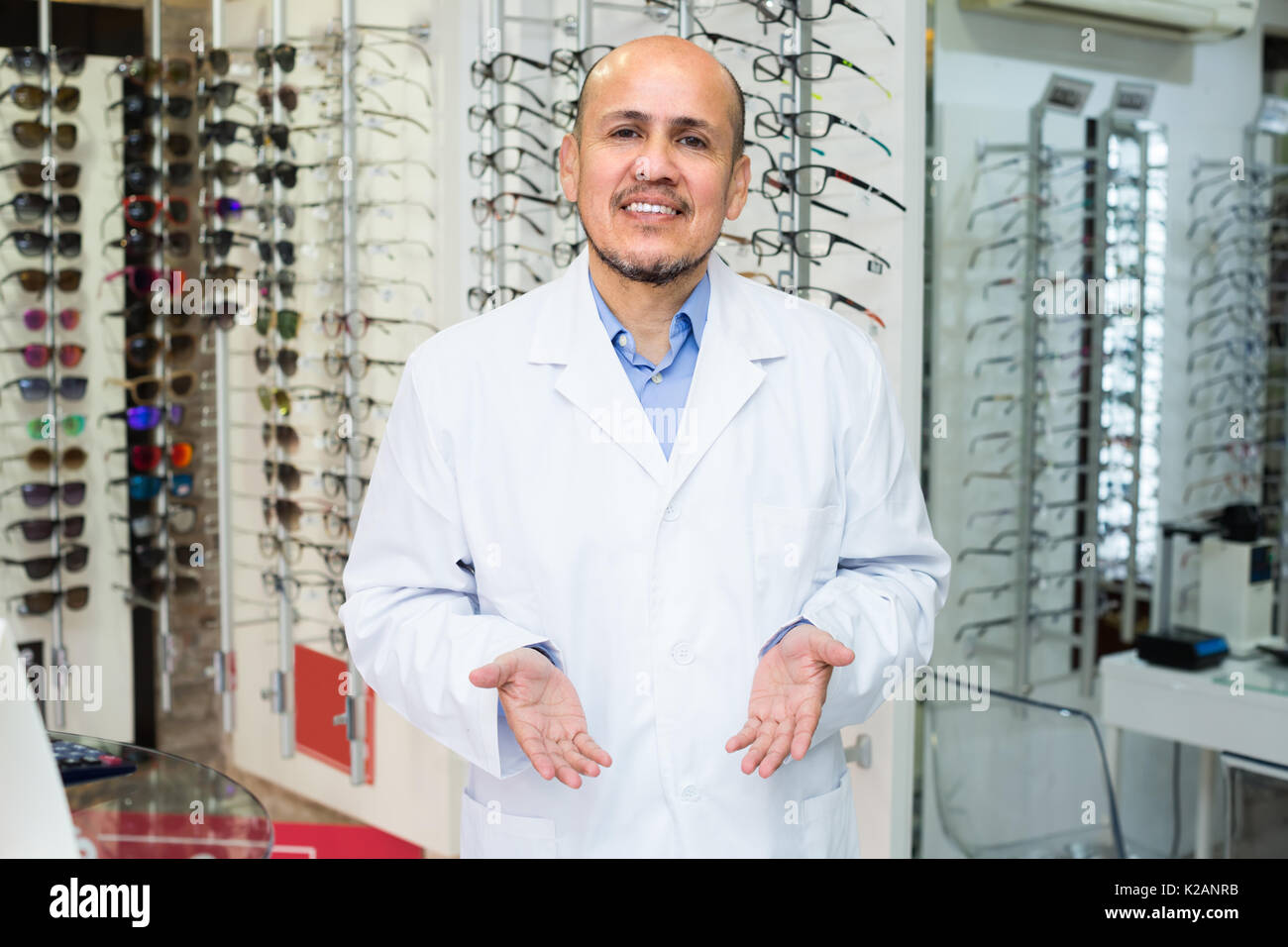 Friendly smiling male optician posing near stand with spectacles and ...