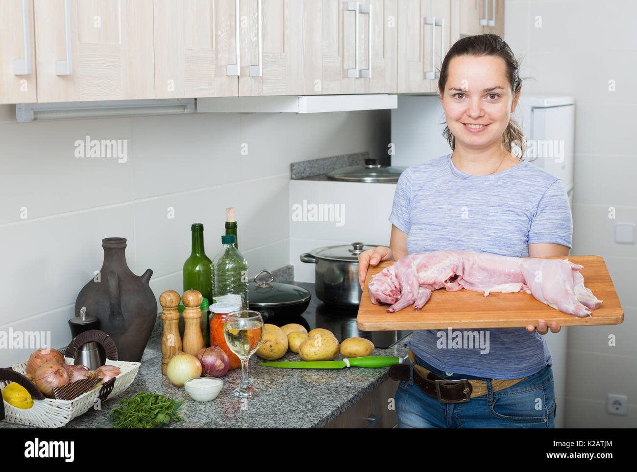 Cheerful young woman preparing whole rabbit to cook on kitchen indoors ...