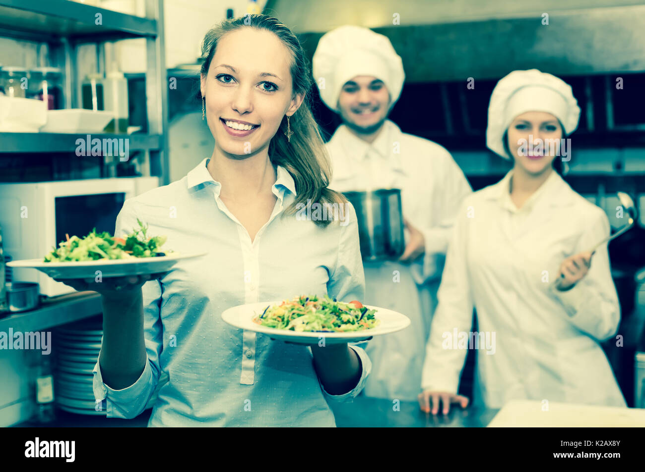 Two chefs and young smiling female waiter at restaurant kitchen ...