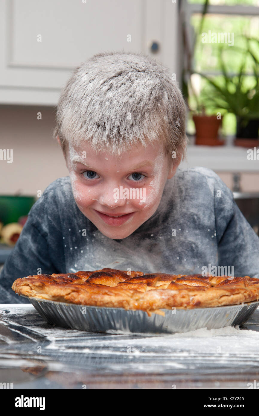 A smiling boy covered in flour and a baked pie on a table in front of ...