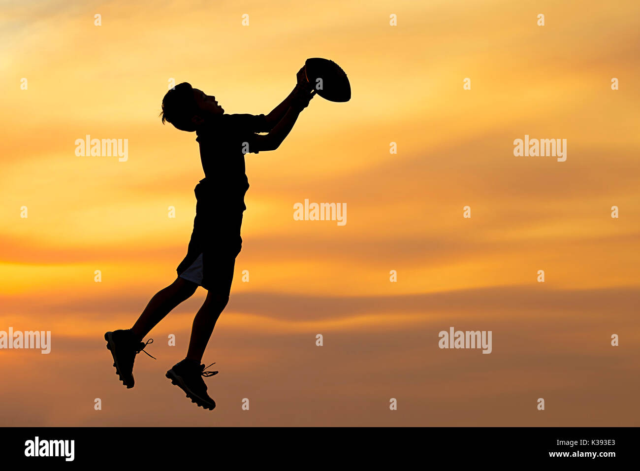 A young boy in silhouette is playing football at sunset against an ...