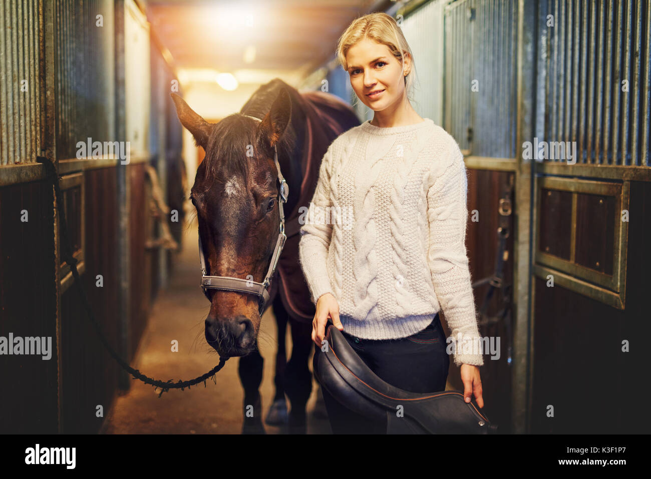 Smiling young woman standing inside a stable holding a saddle while ...