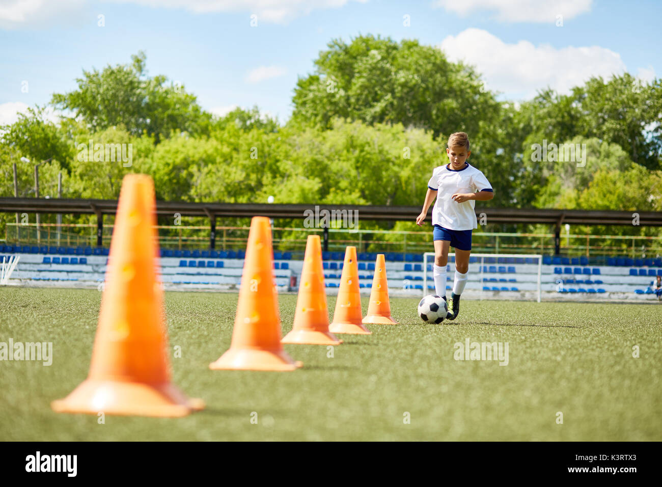Portrait of boy in uniform leading ball between cones during practice ...