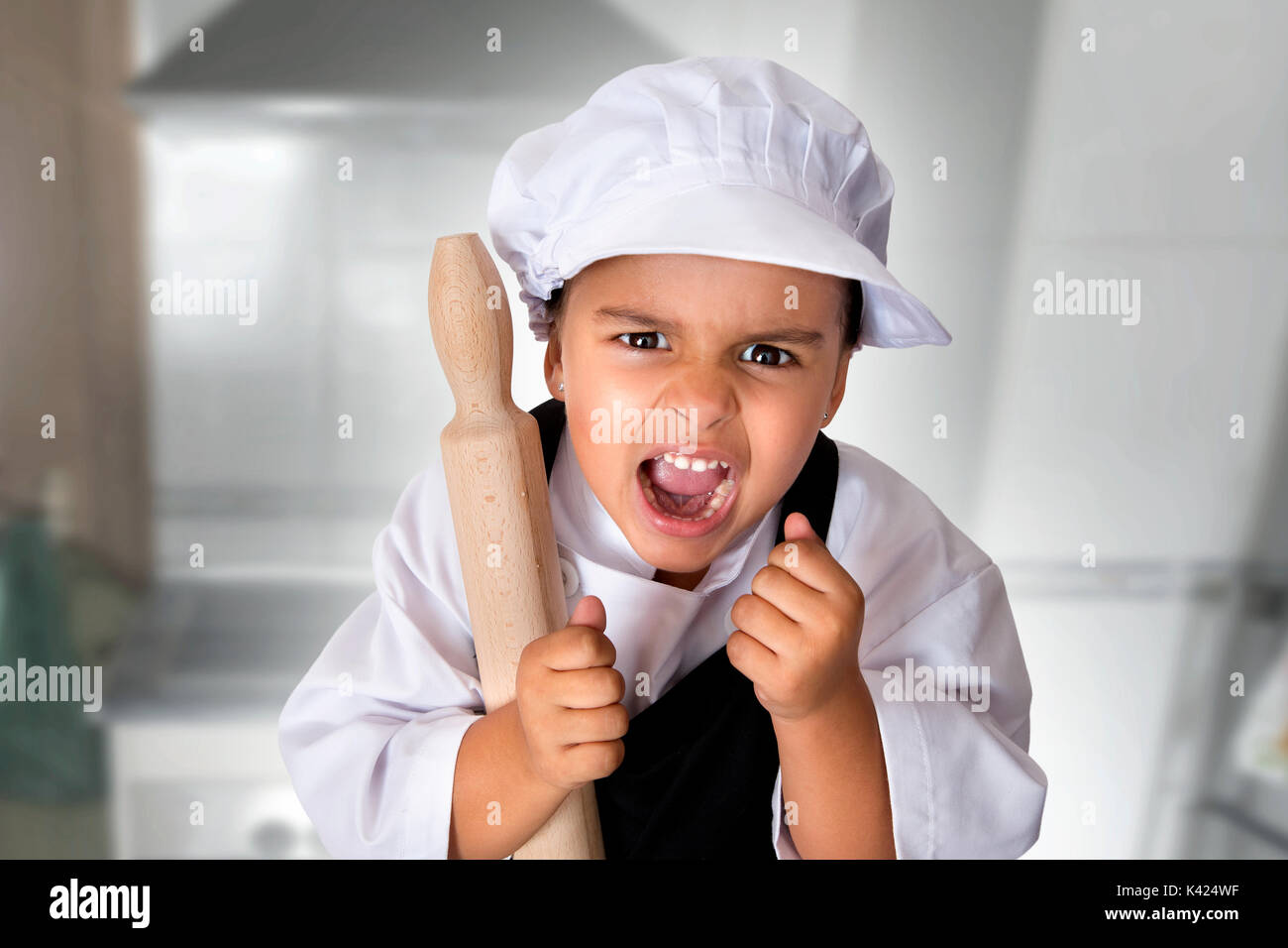 Portrait of a funny four years girl chef with angry face in a kitchen ...