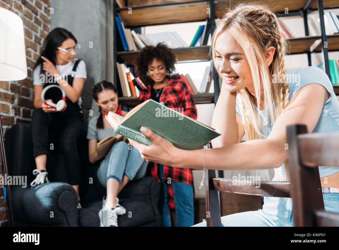 caucasian girl reading book while her friends sitting on sofa at home ...