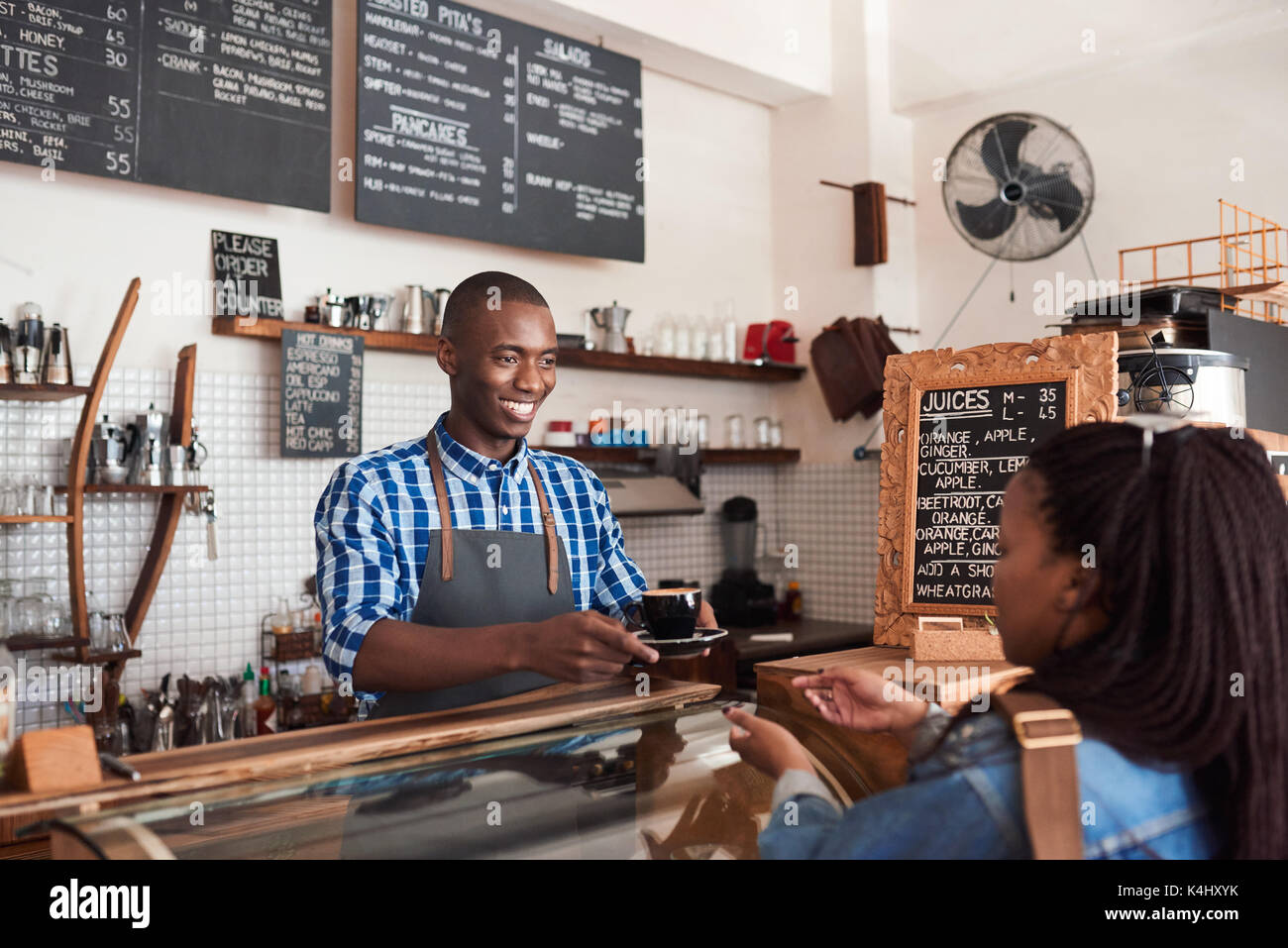 Smiling young African barista standing at the counter of a trendy cafe ...