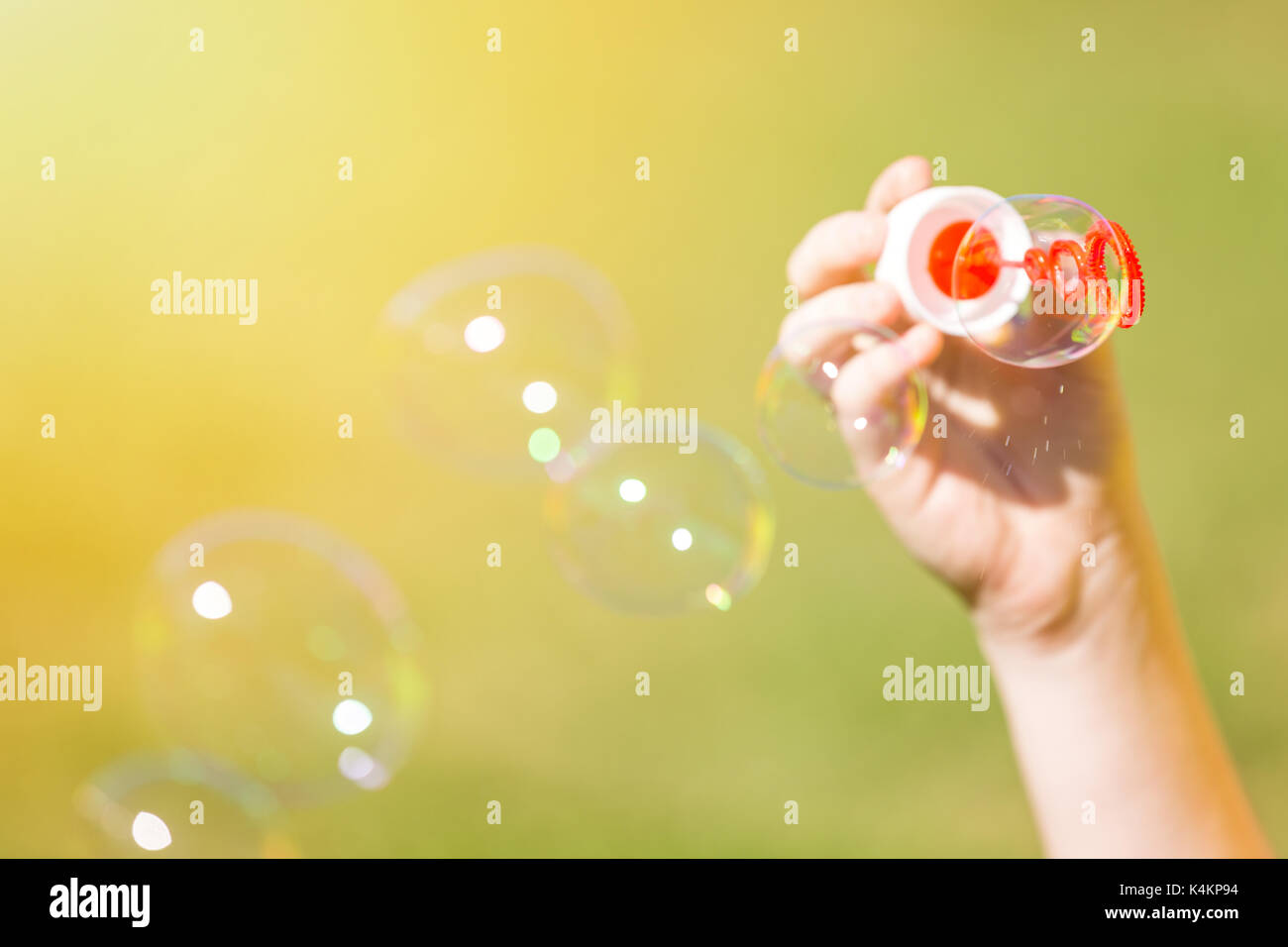 Girls Hand Holding A Bubble Maker And Blowing Soap Bubbles Against The Blurred Grass