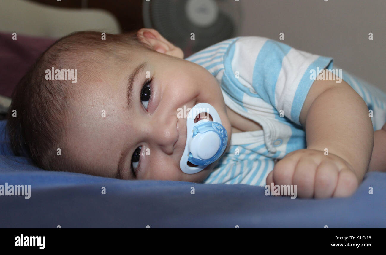 five months old baby boy with dummy laying on bed smilling to camera ...