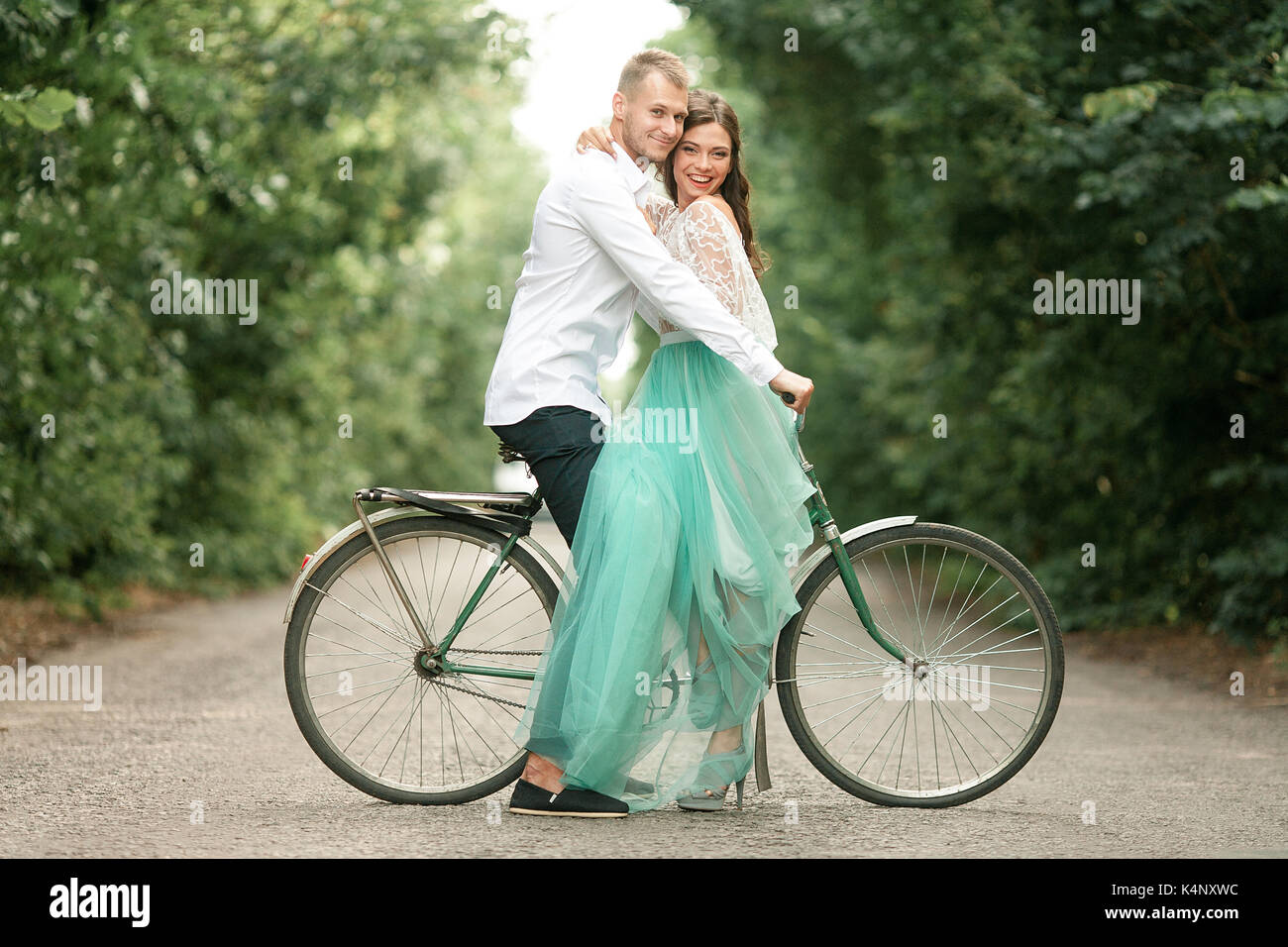 Bride in beautiful wedding dress and groom sit on bicycle on forest ...