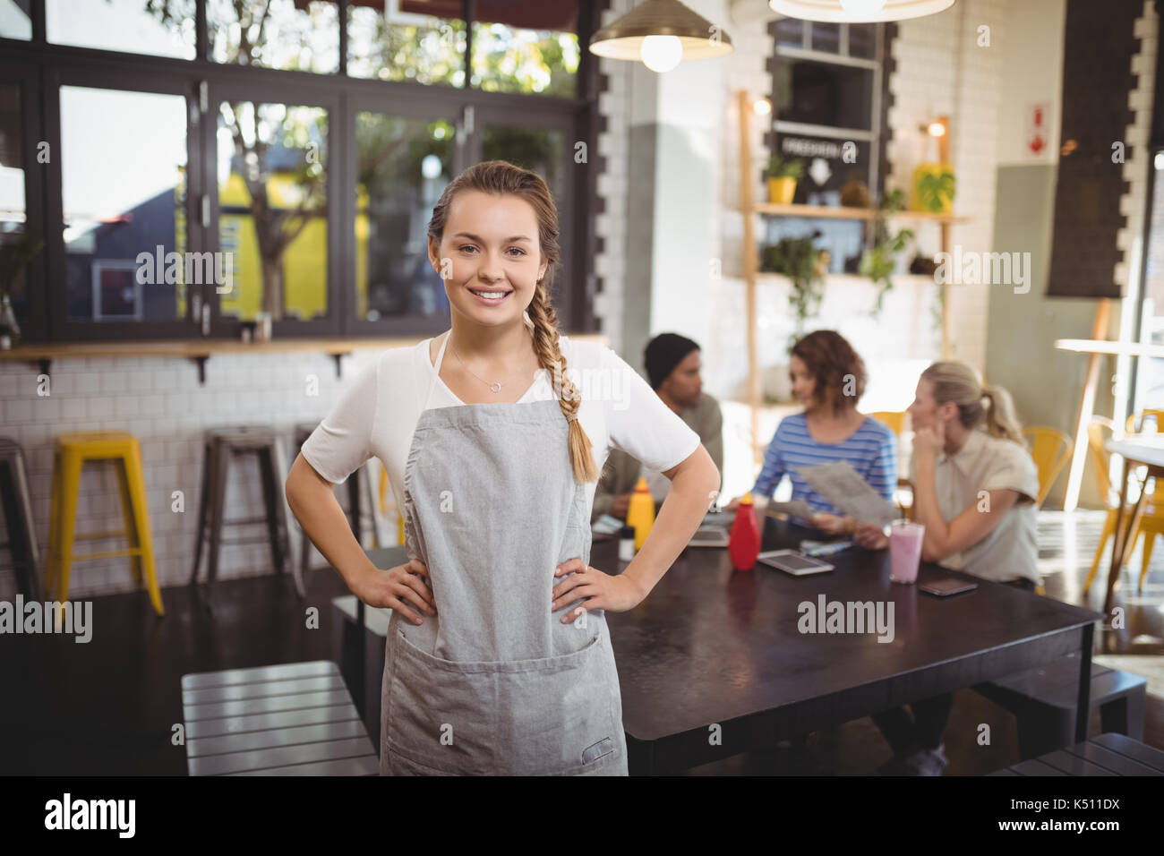 Portrait of smiling young waitress standing with hands on hip at cafe ...