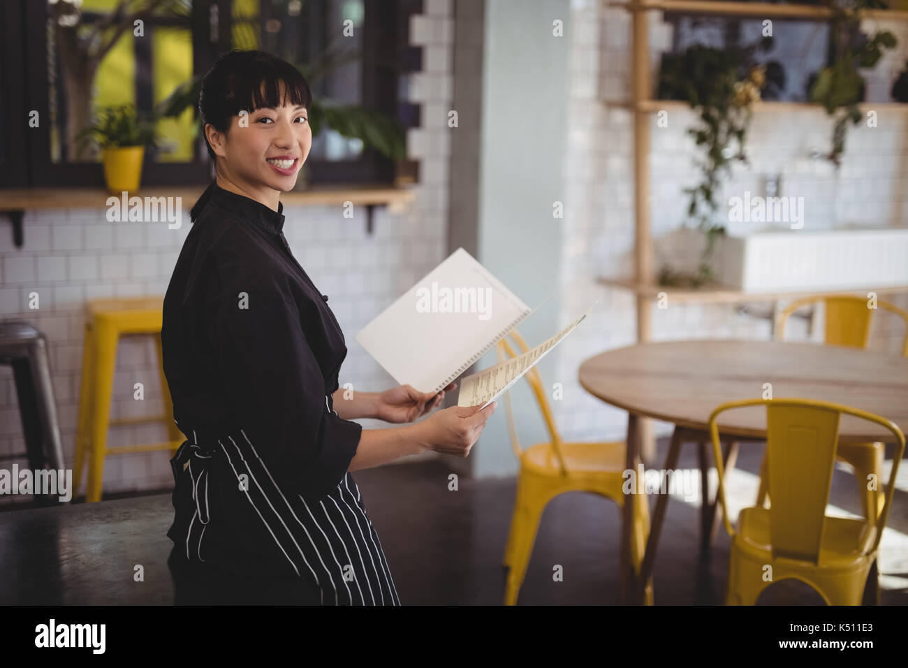 Portrait of young waitress holding menu while leaning on table at ...