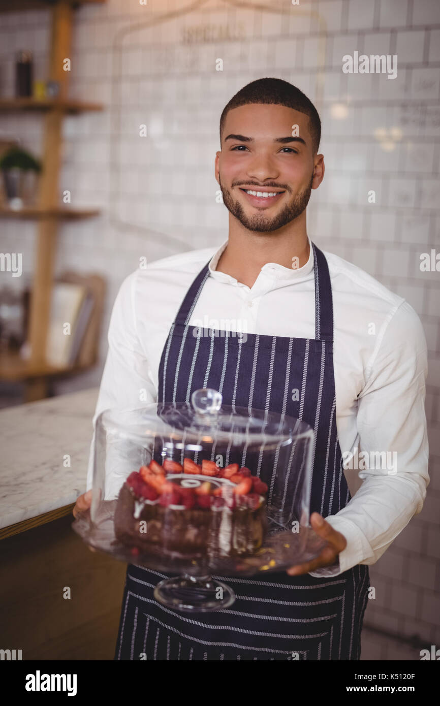 Portrait of smiling young waiter holding cake on glass cakestand at ...